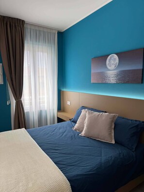 Quarto de hotel com uma cama, um quadro de um mar ao luar e uma janela com cortinas.
