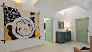 Um corredor com uma grande pintura abstrata, uma porta verde, um armário pequeno e uma cadeira de vime.
