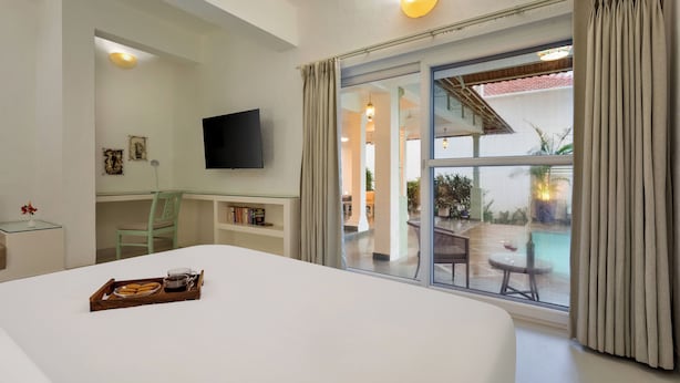 Quarto de hotel com cama, escrivaninha, televisão, estante de livros e vista para área externa com piscina e móveis de pátio.
