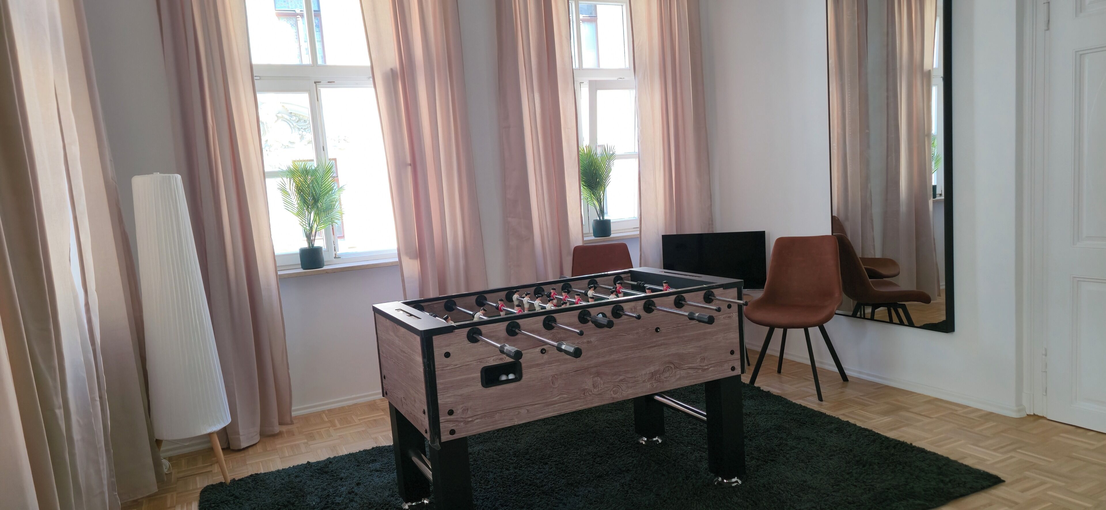 Quarto com mesa de pebolim, uma cadeira, planta em vaso e janela com cortinas.