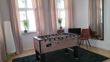 Quarto com mesa de pebolim, uma cadeira, planta em vaso e janela com cortinas.