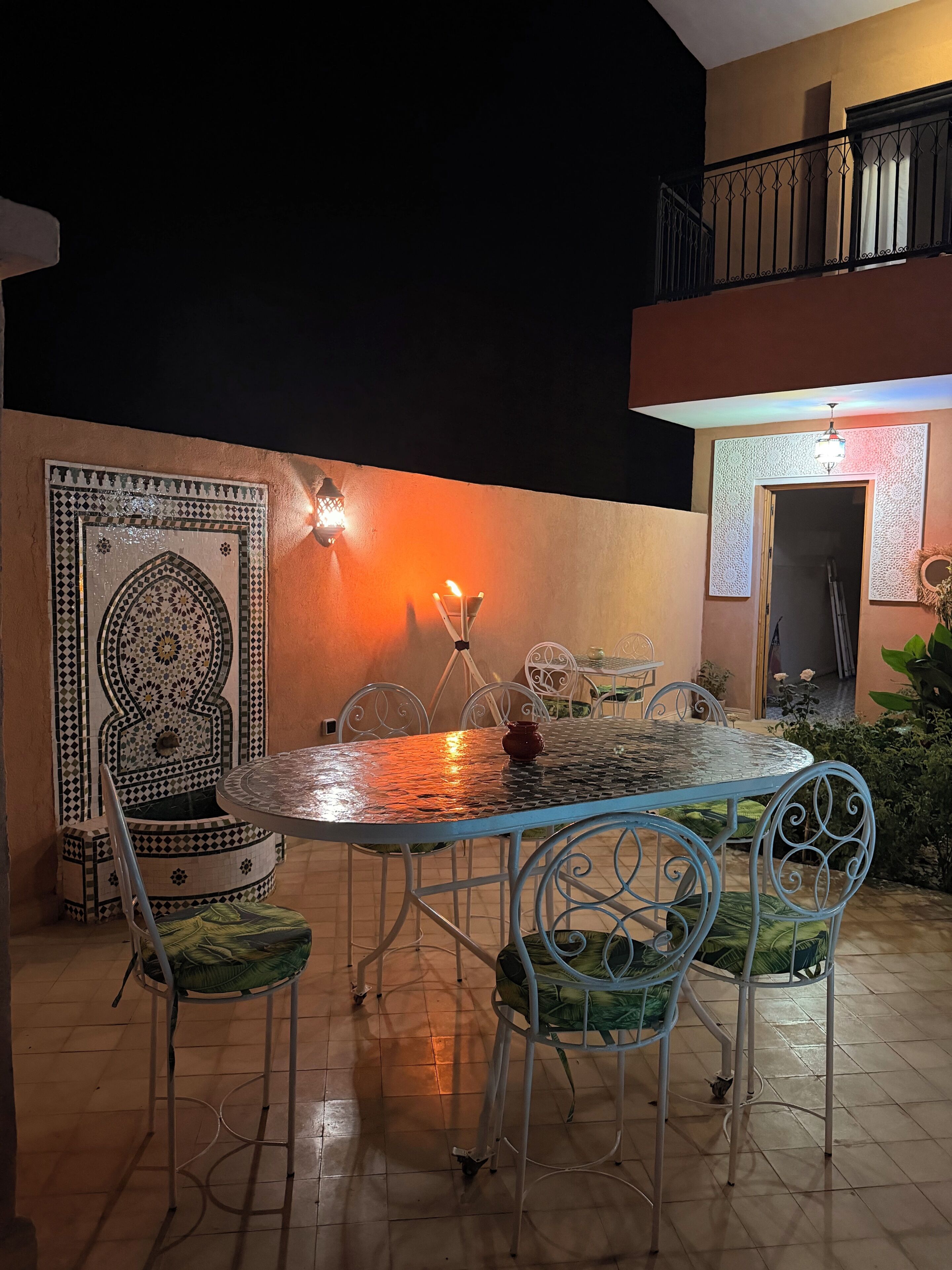 Um pátio externo com mesa redonda e cadeiras, um painel decorativo e uma varanda.