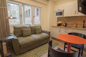 Espaço compacto com sofá, mesa de jantar, área de cozinha e janela com vista para edifícios.