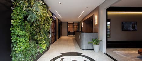 Uma área de recepção de hotel com balcão, um logotipo grande no chão, uma parede com plantas e uma tela de televisão.