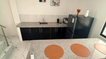 Cozinha compacta com fogão embutido, pia e uma pequena geladeira. Há duas imagens penduradas na parede, uma cadeira e uma mesa com jogo americano.
