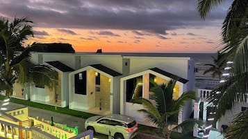 Uma casa moderna de dois andares com um carro estacionado na frente, cercada por palmeiras e vista para o oceano ao pôr do sol.