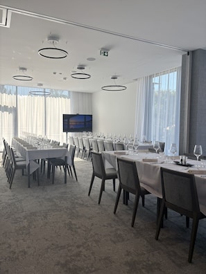 Sala de conferências com várias mesas preparadas para refeições, uma tela de televisão e uma grande janela com cortinas.