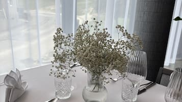 Mesa posta com talheres, copos e um vaso de flores, com vista para a rua através de amplas janelas.