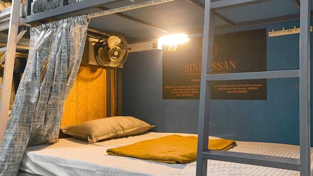 Uma cama de solteiro em beliche com encosto, um ventilador fixado na parede e uma cama com cobertor amarelo.
