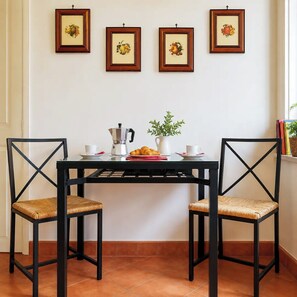 Uma sala de jantar com uma mesa preta, duas cadeiras, uma cafeteira, um vaso com plantas verdes e quatro quadros na parede.
