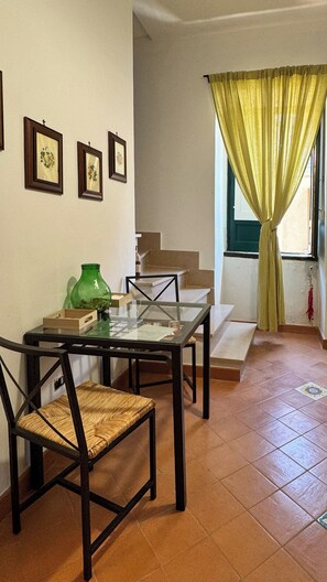 Uma sala de jantar com mesa, cadeiras e um vaso verde. Há quadros nas paredes e uma janela com cortinas amarelas.