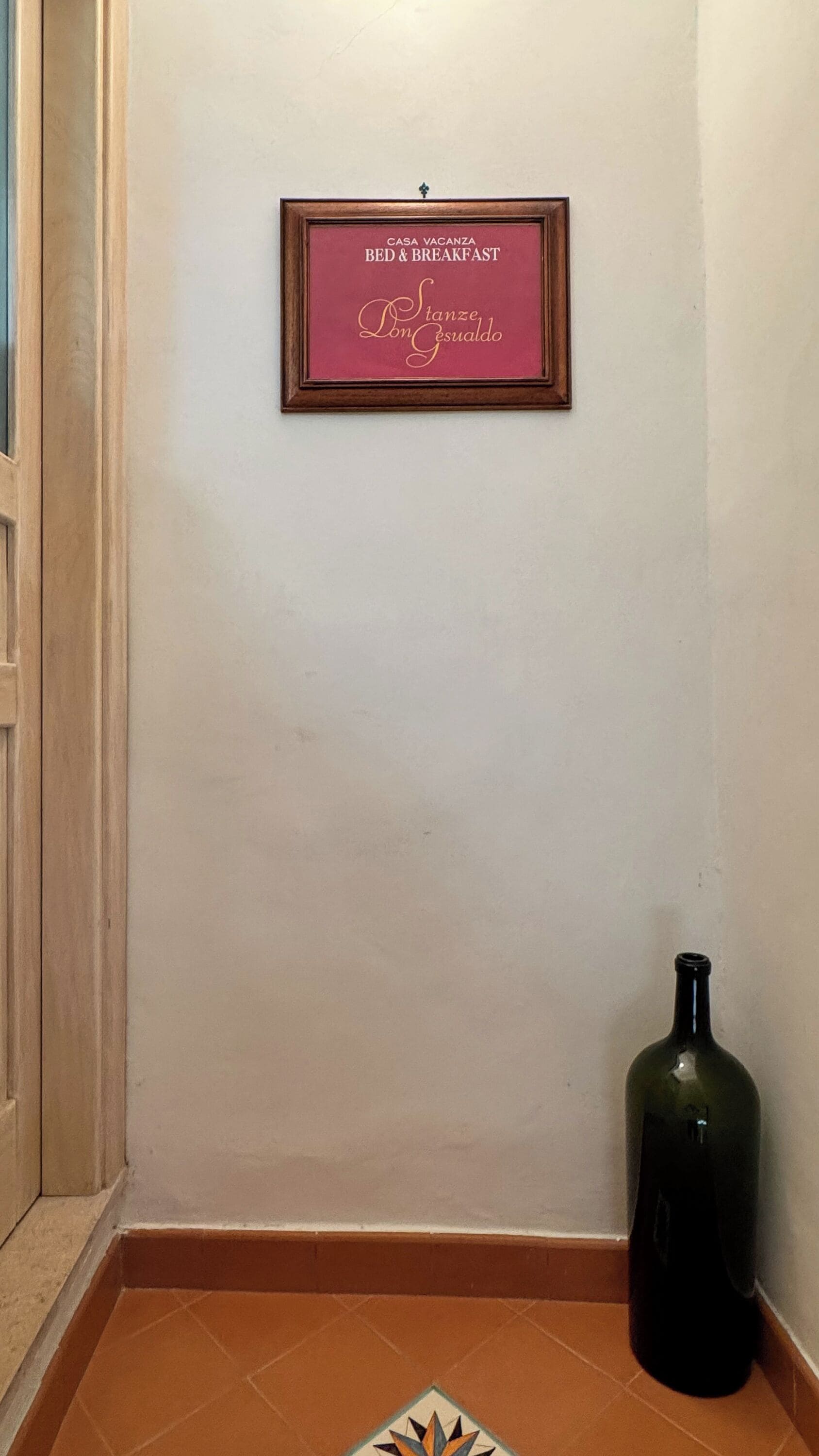 Um letreiro emoldurado na parede, uma garrafa no chão e uma porta à esquerda.
