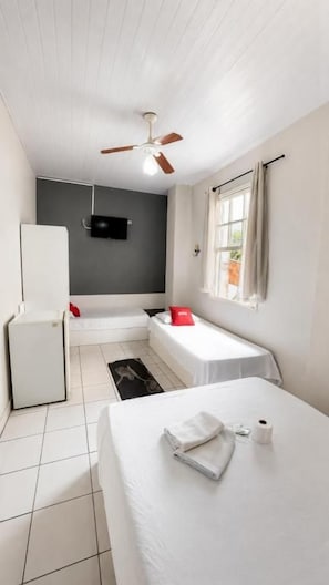 Quarto pequeno e limpo, com cama, escrivaninha, cadeira, ventilador de teto e janela com cortinas.