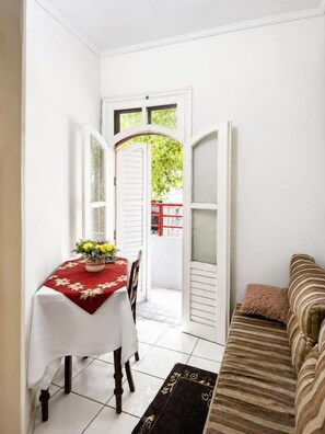 Quarto com mesa de jantar posta com toalha vermelha, uma cadeira e um vaso de flores. Há um sofá com almofada listrada e um tapete marrom. O quarto tem paredes brancas e uma porta que dá para o exterior.