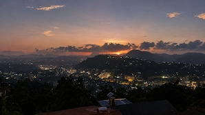 Paisagem urbana noturna com pôr do sol, montanhas ao fundo e vista para uma área residencial.