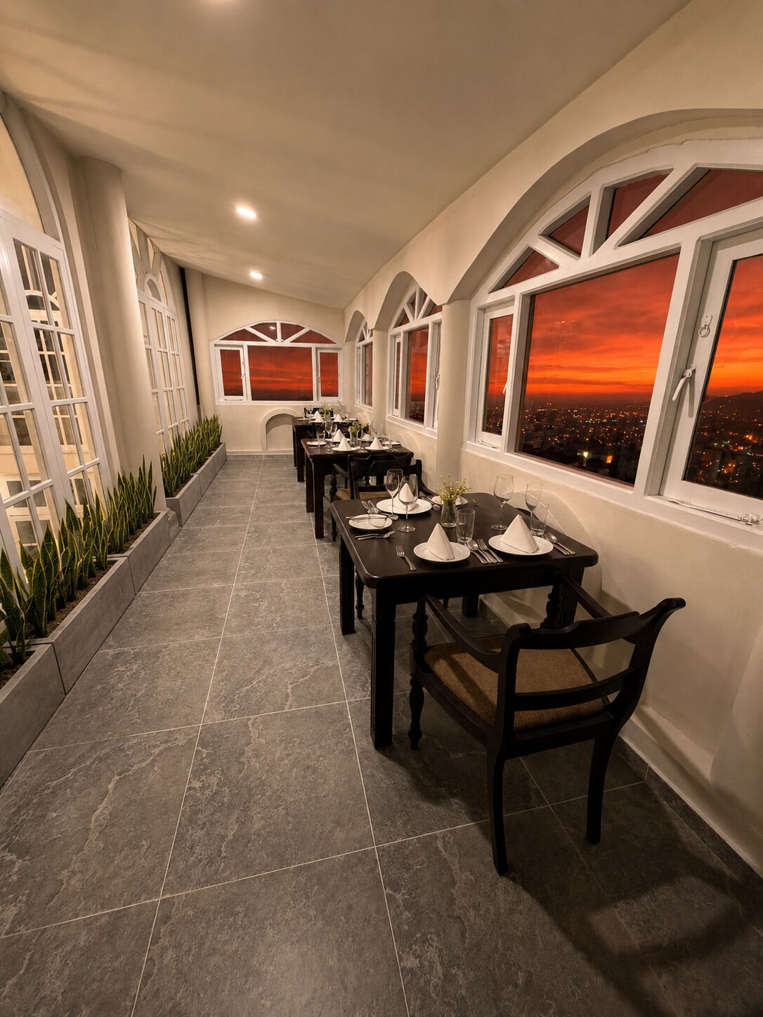 Um corredor com uma longa mesa posta para refeições, grandes janelas com vista para o pôr do sol e piso de azulejos.