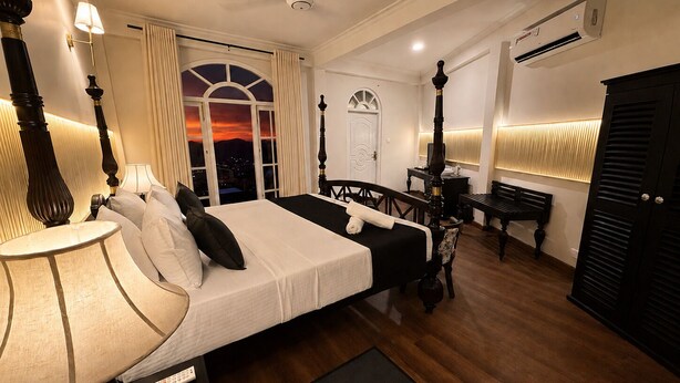 Quarto de hotel com cama, mesa de cabeceira, abajur e vista da cidade ao pôr do sol.