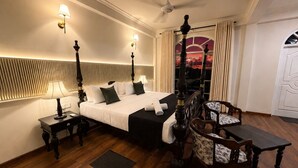 Quarto de hotel com cama de dossel, área de estar com mesa e cadeiras, vista do pôr do sol pela janela e ventilador de teto.