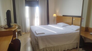 Quarto de hotel com cama, escrivaninha, cadeira, televisão e abajur.