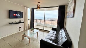 Sala de estar com sofá de couro preto, mesa de centro de vidro, televisão de tela plana na parede e vista para o mar através de grandes janelas.