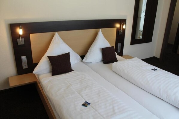 Quarto de hotel com duas camas, cada uma com roupa de cama branca e almofadas castanhas, cabeceira de madeira, luminárias de cabeceira e um espelho na parede.