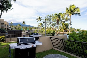 Um pátio com duas churrasqueiras, uma mesa e um corrimão com vista para um jardim com palmeiras e montanhas.