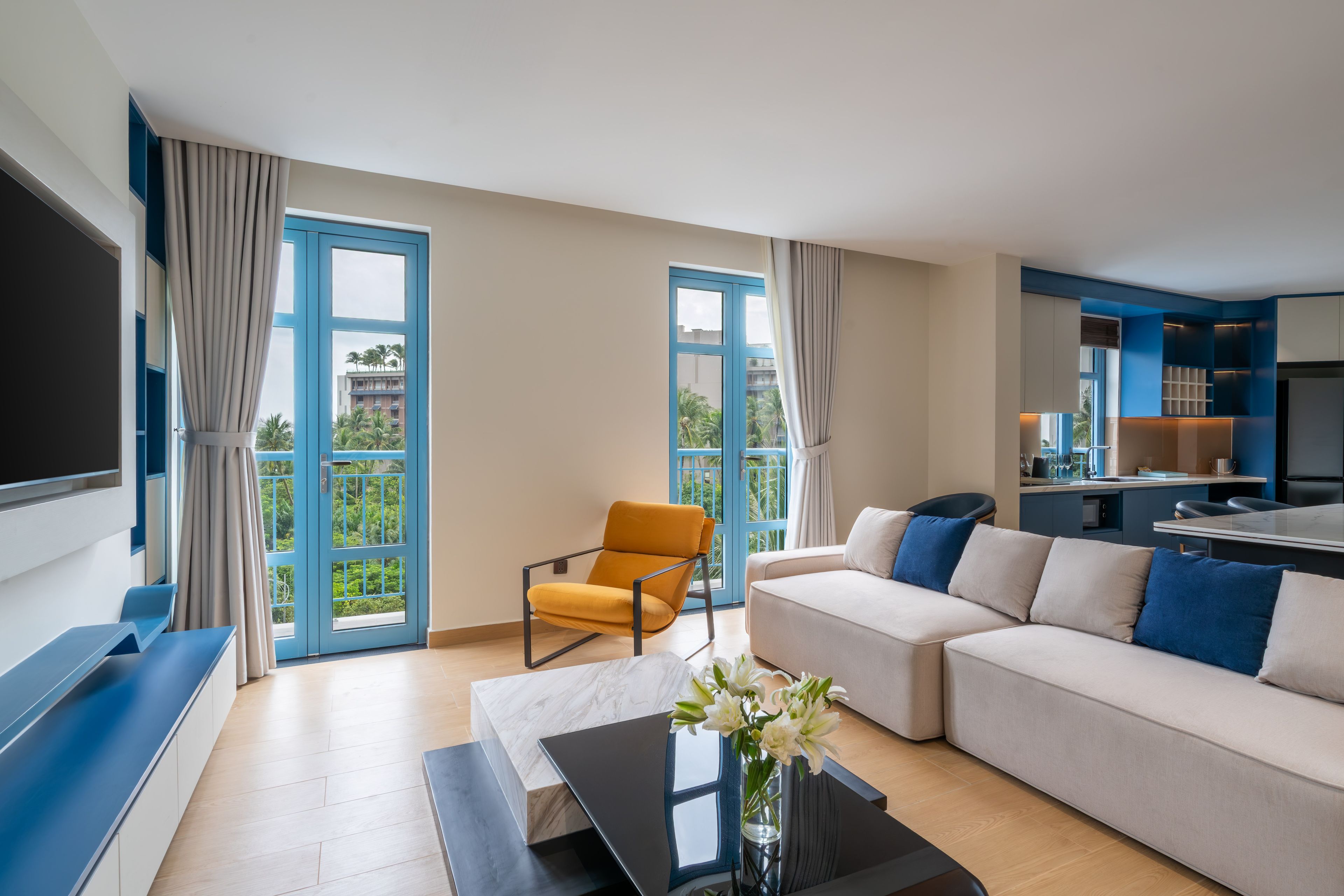 Uma sala de estar moderna com sofá, mesa de centro, televisão e uma poltrona. Há grandes janelas com cortinas, uma área de cozinha e uma varanda com vista para palmeiras.