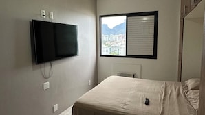 Quarto com cama, ventilador de teto, televisão fixada na parede e janela com vista para edifícios.