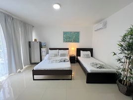 Quarto de hotel com uma cama grande, uma cama menor, geladeira, bebedouro, luminária de cabeceira, ar-condicionado fixado na parede e um vaso de plantas.