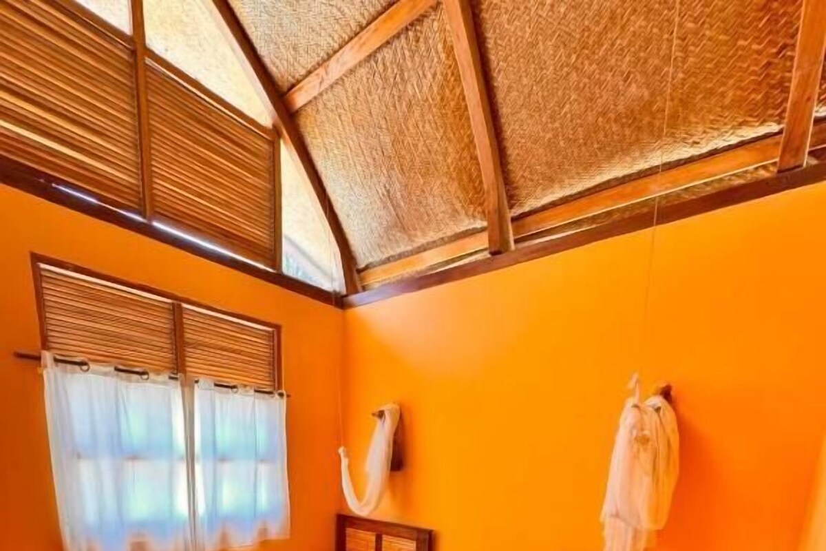 Quarto com duas camas de solteiro, piso de madeira, telhado de palha e janela com cortinas.