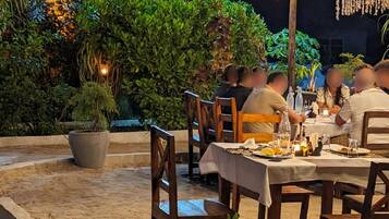 Uma área de refeições ao ar livre com mesas e cadeiras, rodeada por vegetação e luzes de fada.