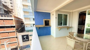 Uma varanda com mesa de vidro e cadeiras de vime, parede com azulejos azuis e vista para um edifício em construção.
