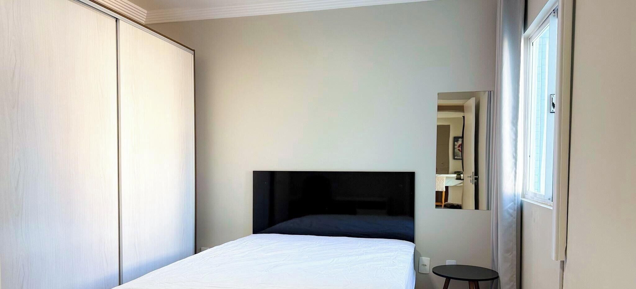 Quarto com cama, mesa de cabeceira, guarda-roupa e janela.