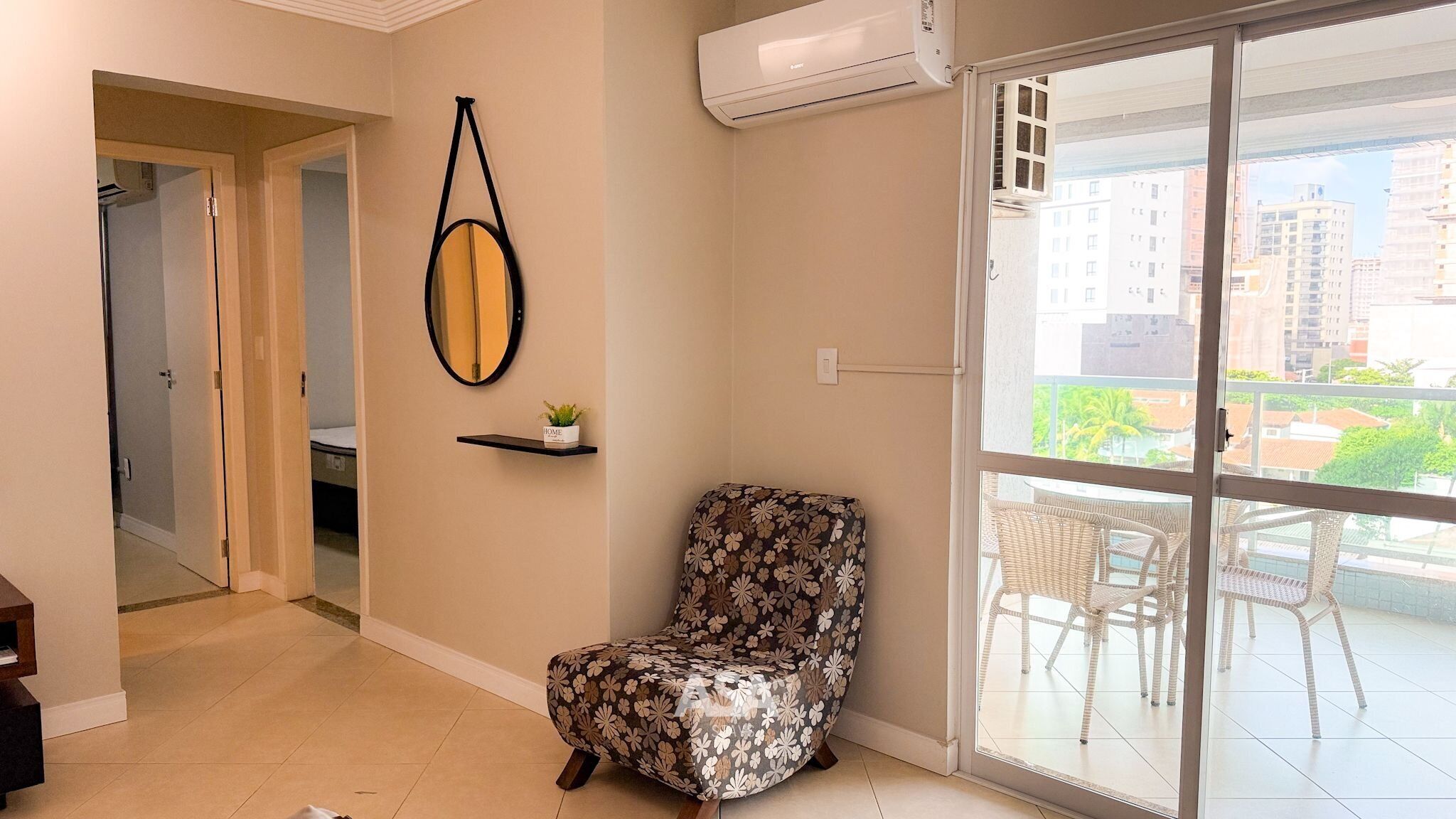 Uma sala de estar com uma poltrona, um espelho fixado na parede, um pequeno estante com uma planta e uma porta de vidro deslizante que dá para uma área externa com móveis de vime.
