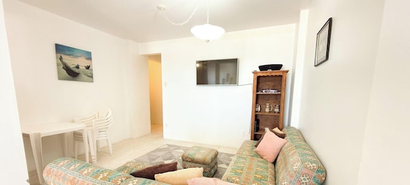 Uma sala de estar com sofá, mesa de jantar com cadeiras, televisão e estante de livros.