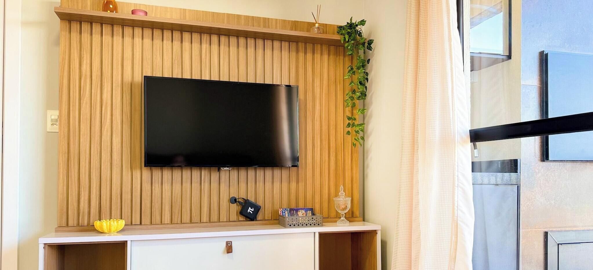 Uma sala de estar moderna com uma televisão de tela plana presa a uma parede de madeira, uma pequena planta e um recipiente de vidro sobre uma mesa de console branca.