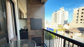 Varanda com mesa de vidro e uma cadeira, pia pequena e vista para edifícios ao redor.