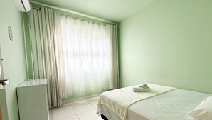Quarto com cama, janela com cortinas, radiador e ar condicionado fixado na parede.