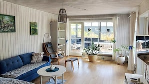 Sala de estar com sofá azul, mesa de centro branca, estante de livros e uma grande janela com vista para o exterior.