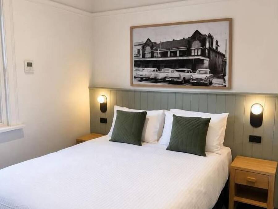 Quarto de hotel com uma cama grande, duas mesas de cabeceira, luminária fixada na parede e uma moldura com imagem sobre a cama.