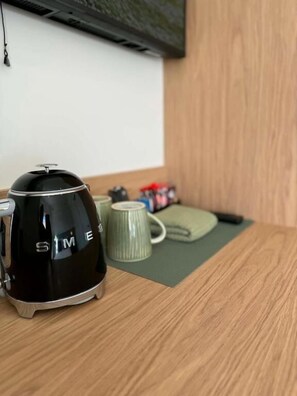 Uma cafeteira preta sobre uma mesa de madeira com um tapete verde, uma caneca branca e uma almofada verde.