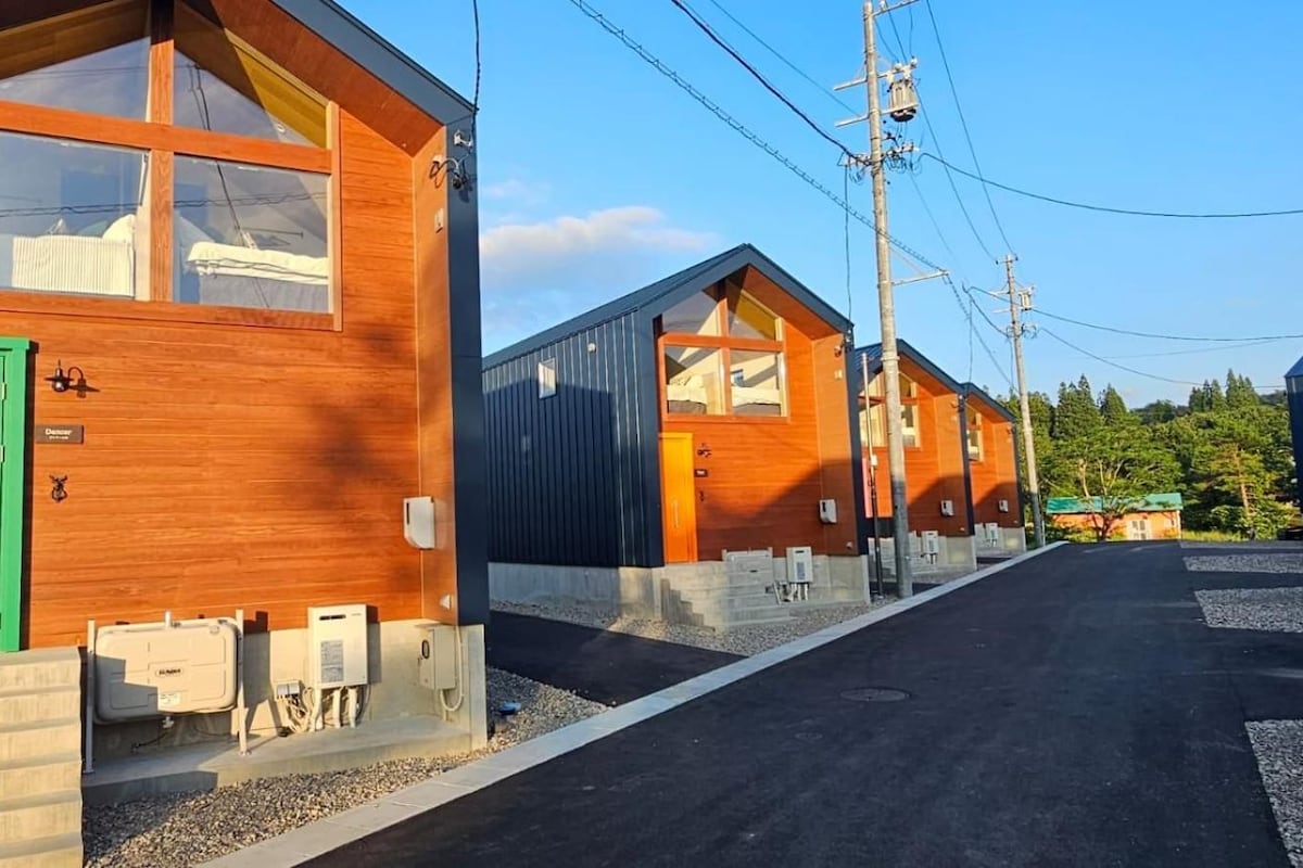 Fileira de cabanas com fachada de madeira, uma porta verde e caixas de eletricidade externas.