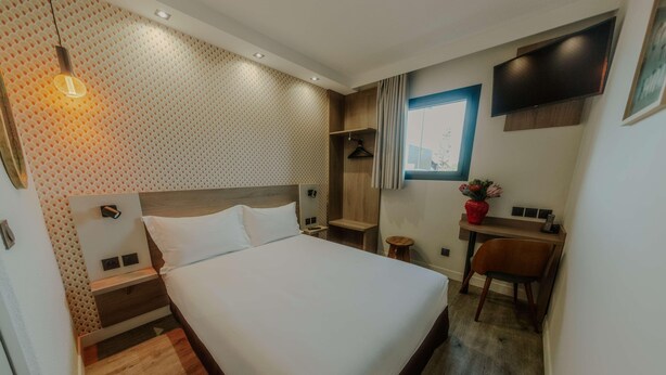 Quarto de hotel moderno com cama grande, mesas de cabeceira, escrivaninha e televisão na parede.