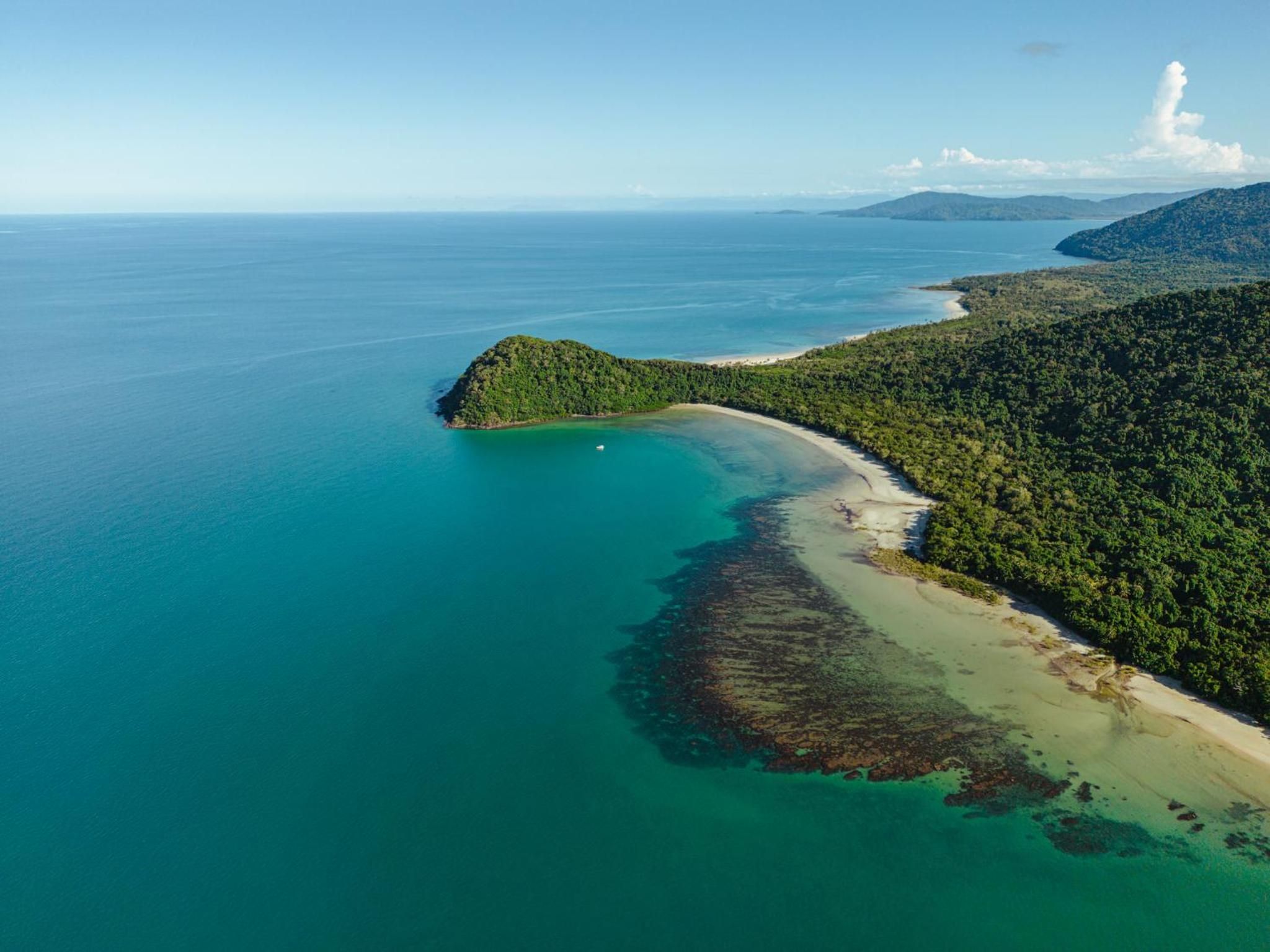 Vista aérea de uma área costeira com praia, águas cristalinas e uma península arborizada.