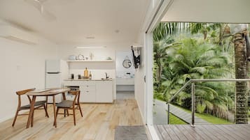 Cozinha moderna com armários brancos, sala de jantar com cadeiras de madeira e vista para um jardim tropical exuberante através de uma porta de vidro.