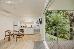 Cozinha moderna com armários brancos, sala de jantar com cadeiras de madeira e vista para um jardim tropical exuberante através de uma porta de vidro.