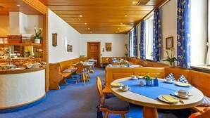Espaço de refeições de hotel com mesas de madeira, toalhas de mesa azuis e cozinha visível ao fundo.