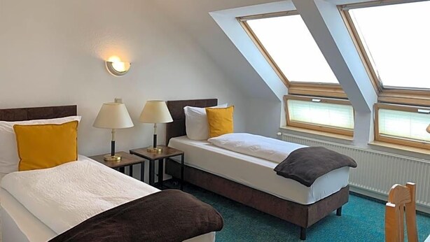 Quarto de hotel com duas camas, claraboia, mesa de cabeceira com luminárias e vista para o exterior.