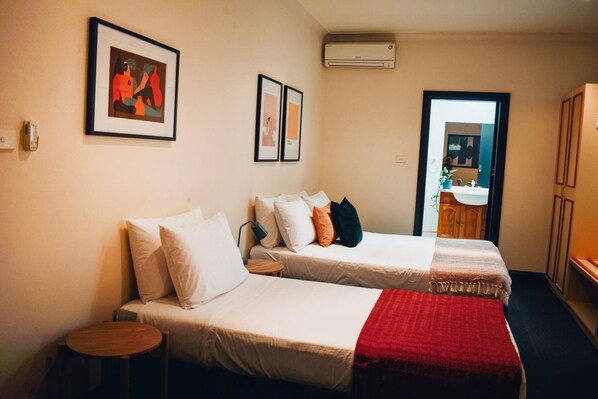Quarto de hotel com duas camas, uma mesinha, um cobertor vermelho e uma porta que dá para um banheiro.