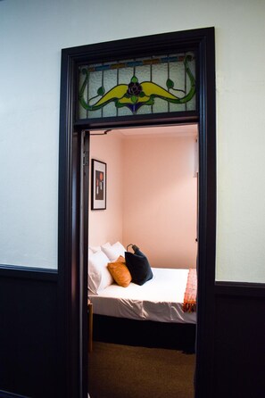 Uma porta estreita dá acesso a um pequeno quarto com uma cama, um quadro na parede e um vitral decorativo sobre a porta.
